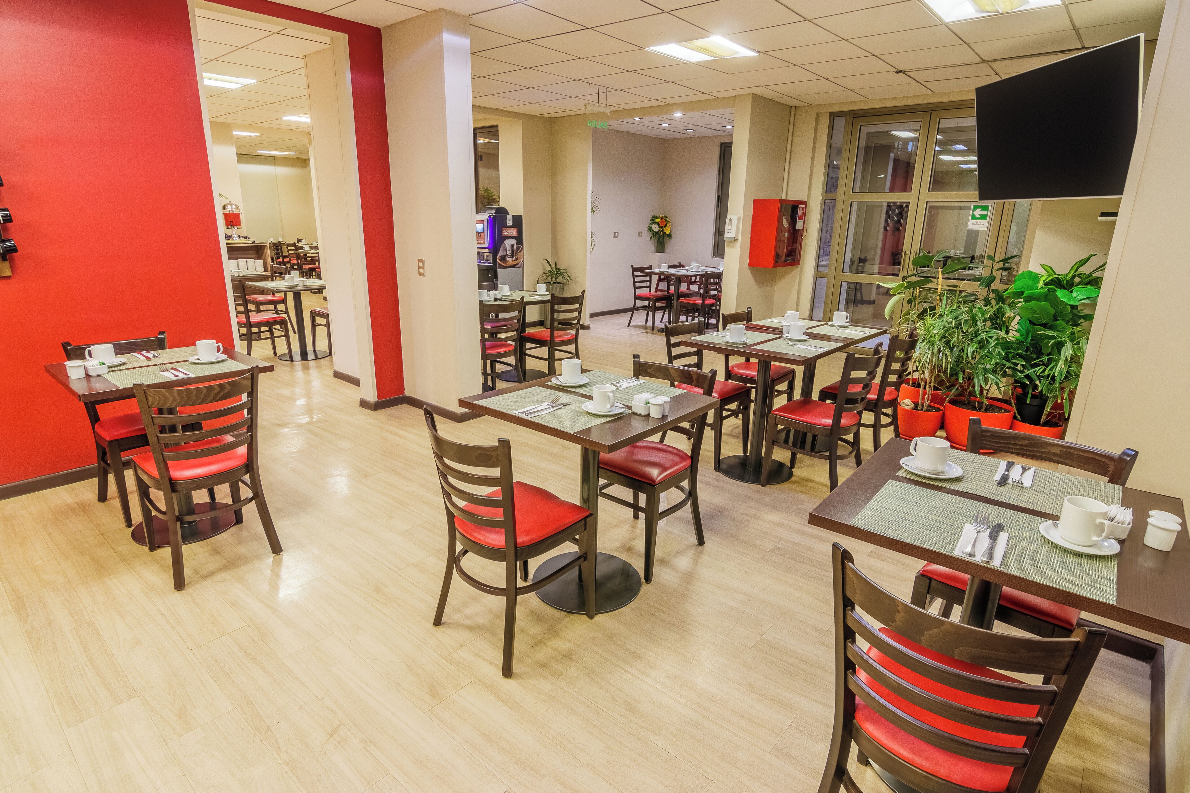 Uma sala de jantar de hotel com várias mesas e cadeiras, uma máquina de café e plantas em vasos.