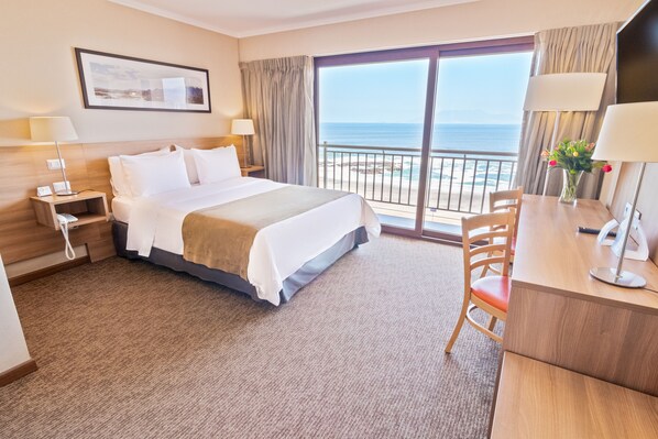Quarto de hotel com cama, escrivaninha com cadeira, telefone e vista para o oceano.