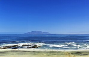 Uma praia com águas azul-claras, costa rochosa e montanhas ao longe.