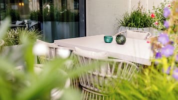Uma área de estar ao ar livre com uma mesa branca e cadeiras de vime, cercada por plantas e flores.