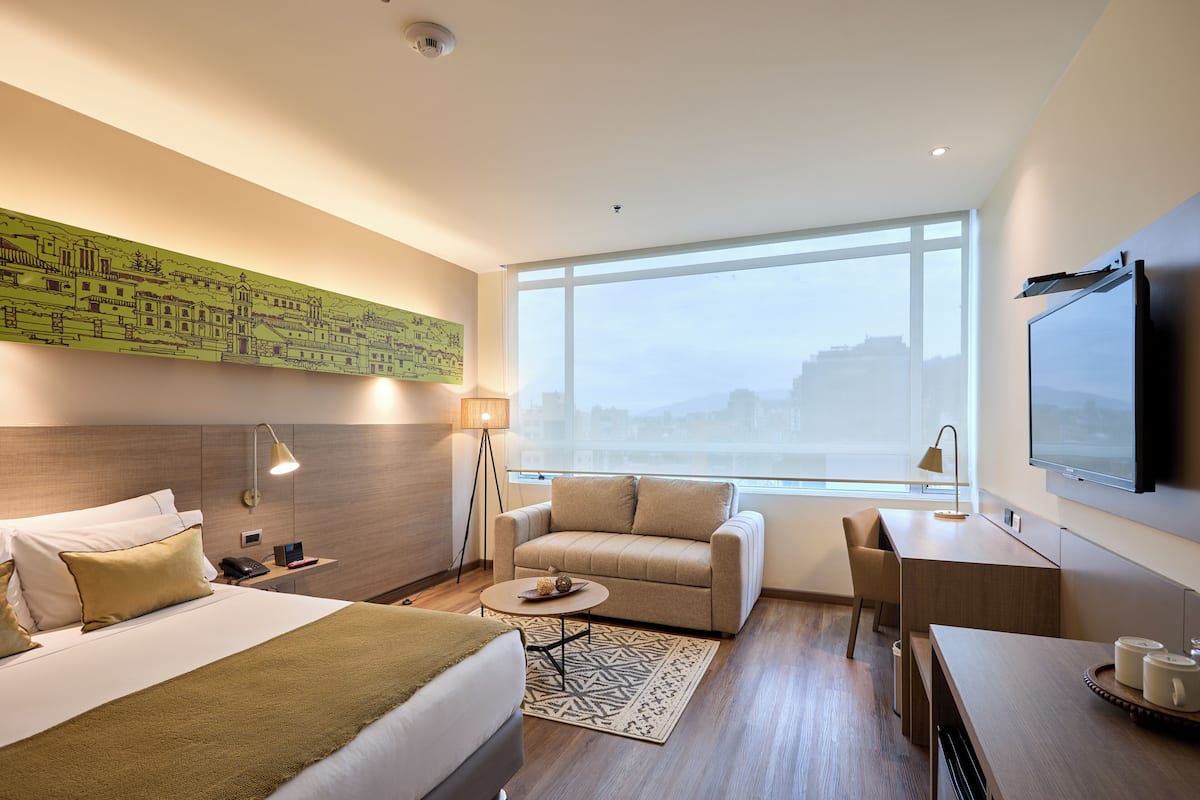 Quarto de hotel com cama, sofá, escrivaninha, televisão e uma janela grande com vista.