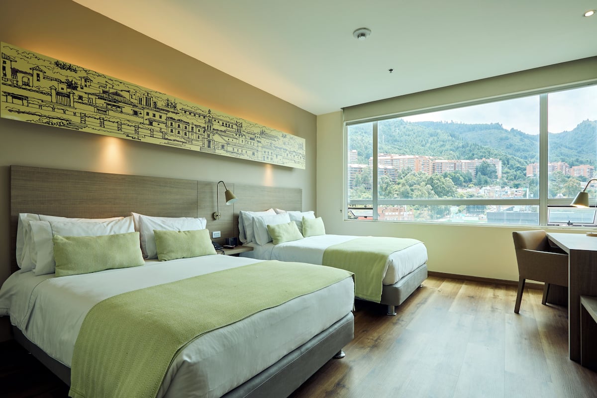 Quarto de hotel com duas camas, uma escrivaninha, uma cadeira, uma janela grande com vista para edifícios e colinas, e uma obra de arte na parede.