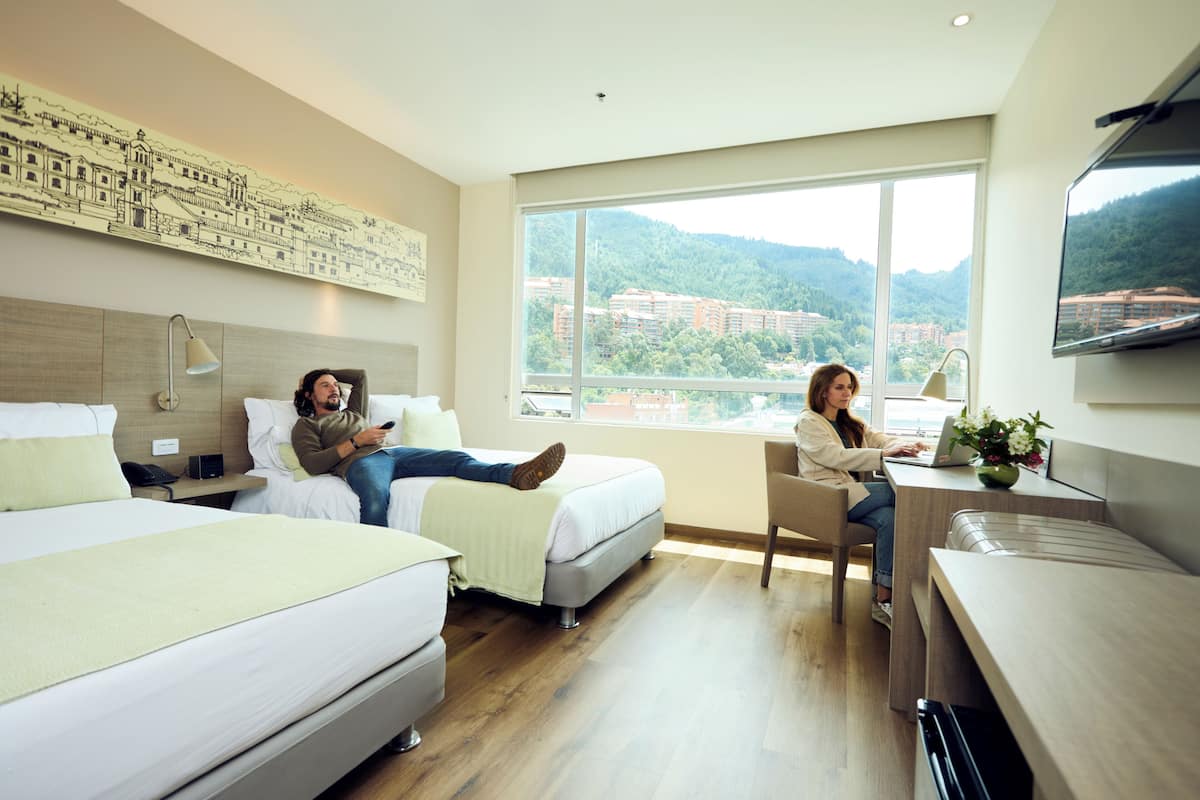 Quarto de hotel com duas camas, uma escrivaninha e uma janela grande com vista para edifícios e colinas.