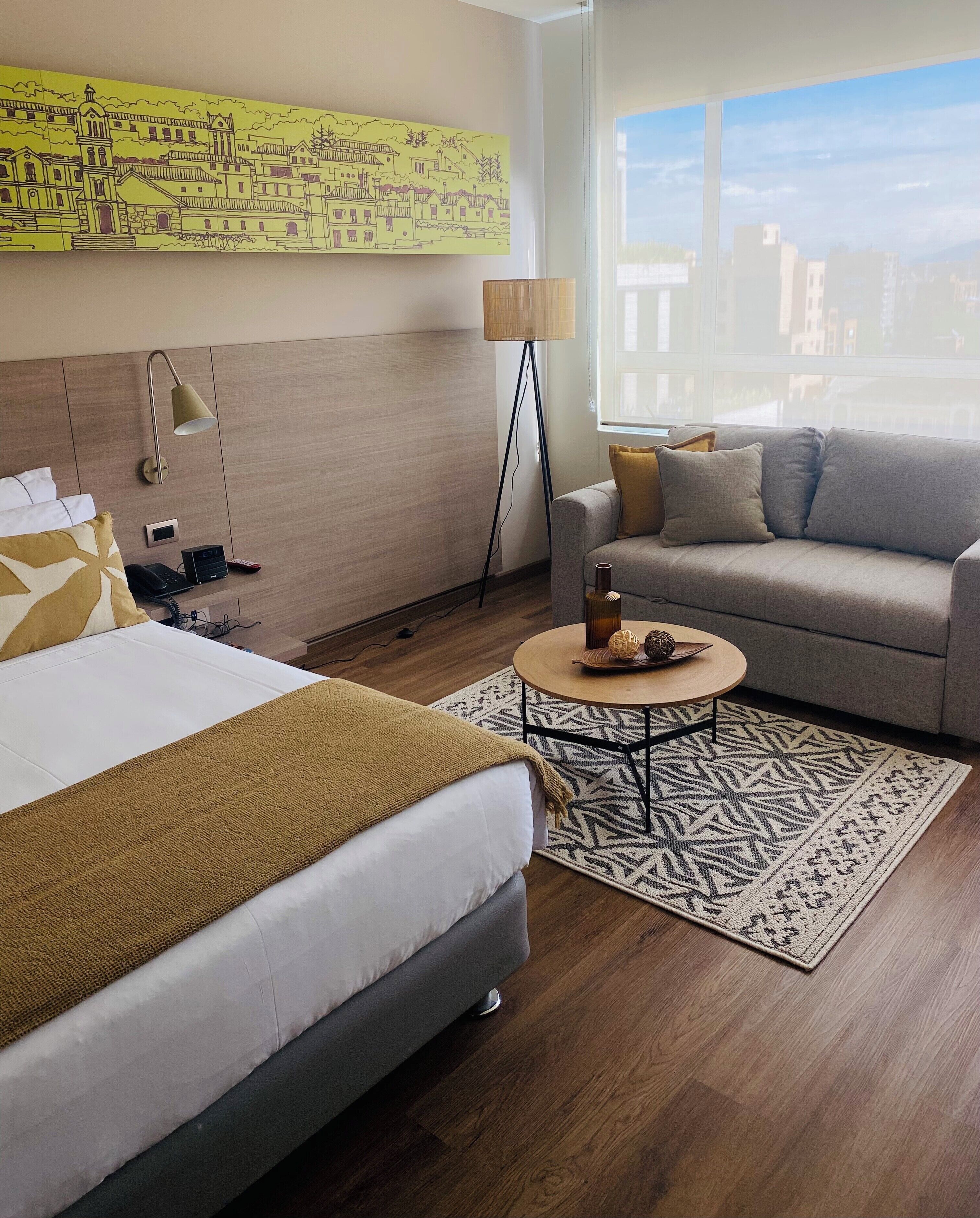 Quarto de hotel com cama, sofá, mesa de centro, luminária, quadro e vista da cidade.