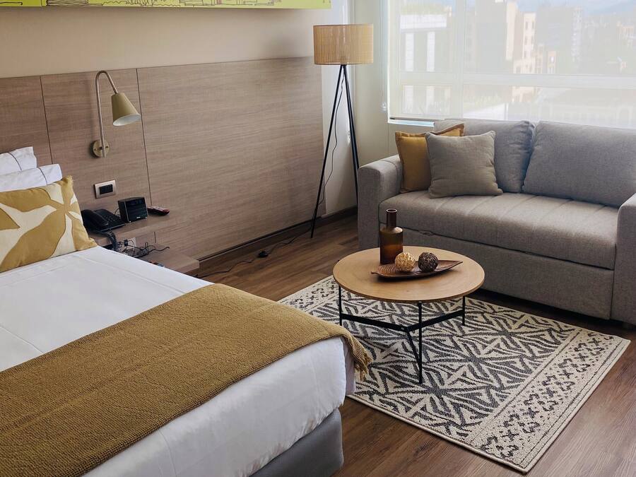 Quarto de hotel com cama, sofá, mesa de centro, luminária, quadro e vista da cidade.