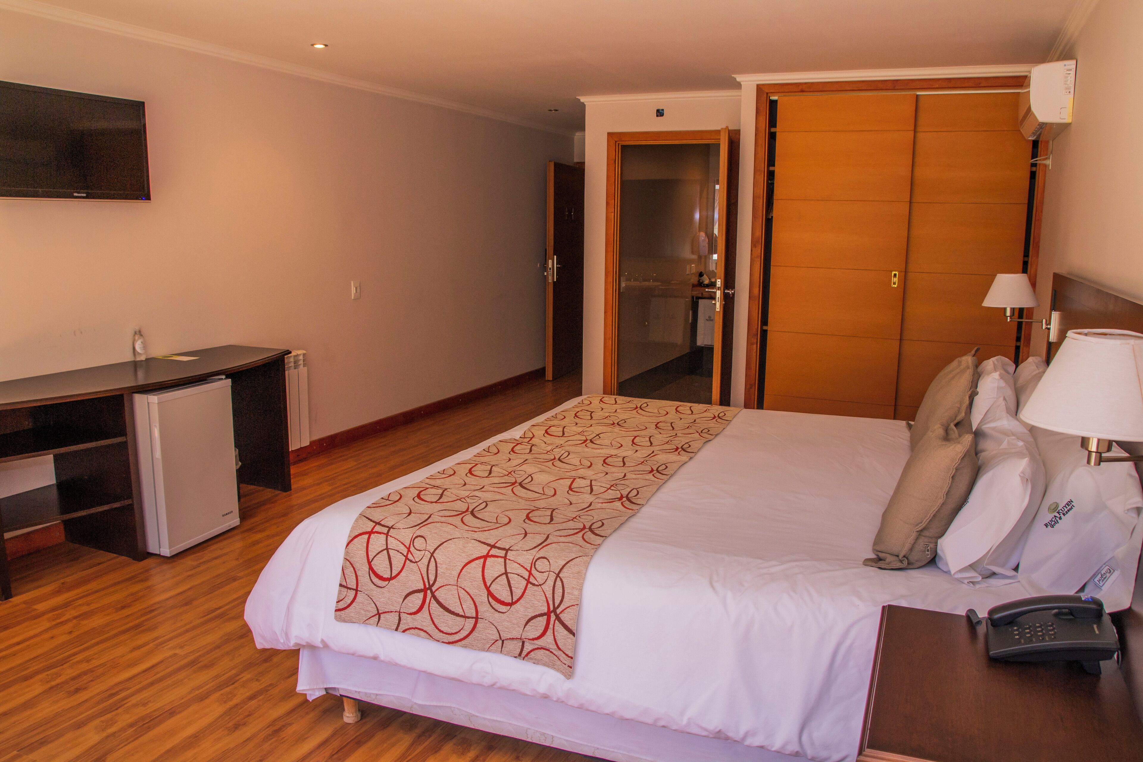 Quarto de hotel com cama, mesa de cabeceira, luminária, telefone e escrivaninha com computador.