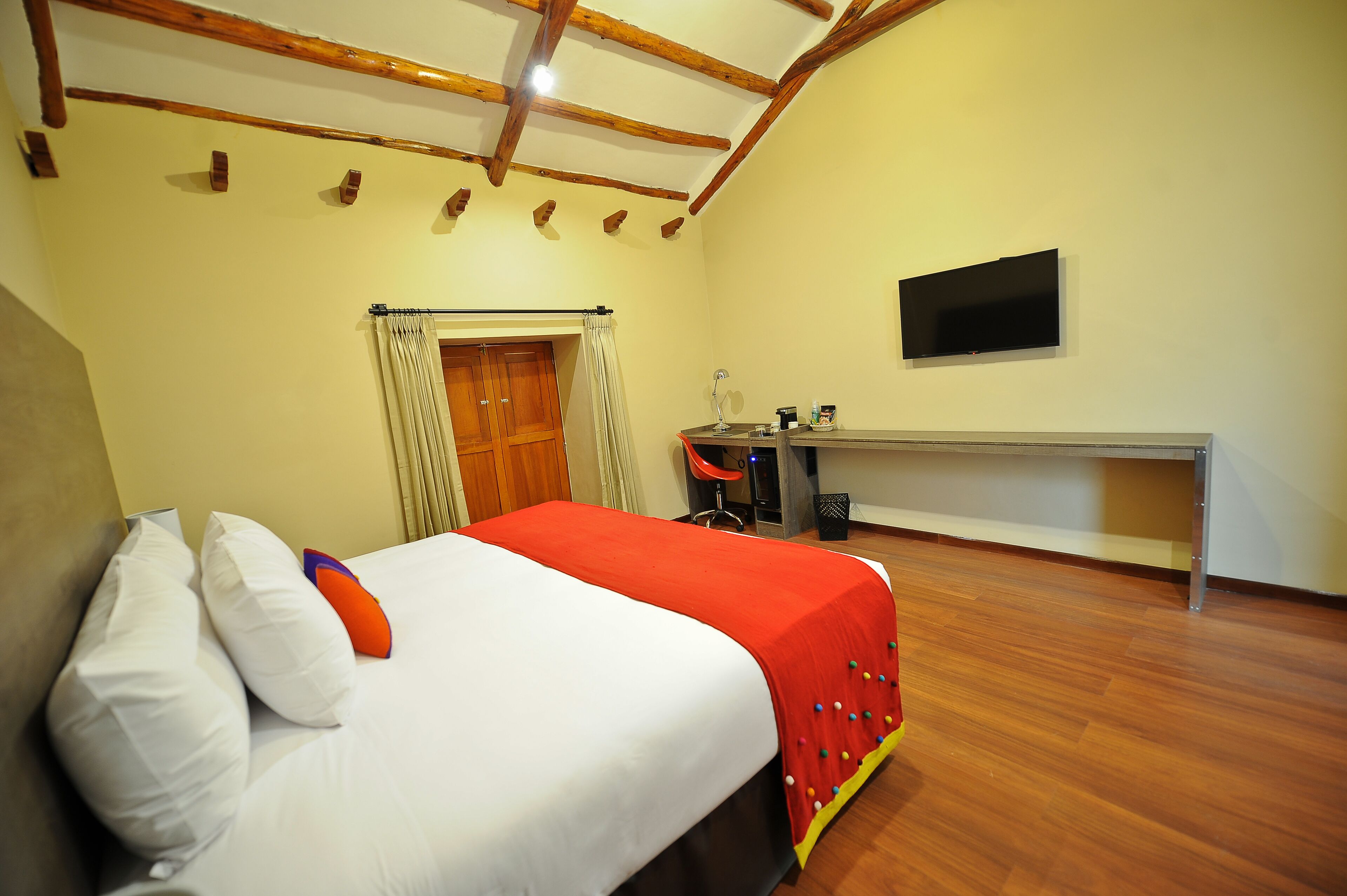 Quarto de hotel com cama, escrivaninha, televisão e teto de madeira.