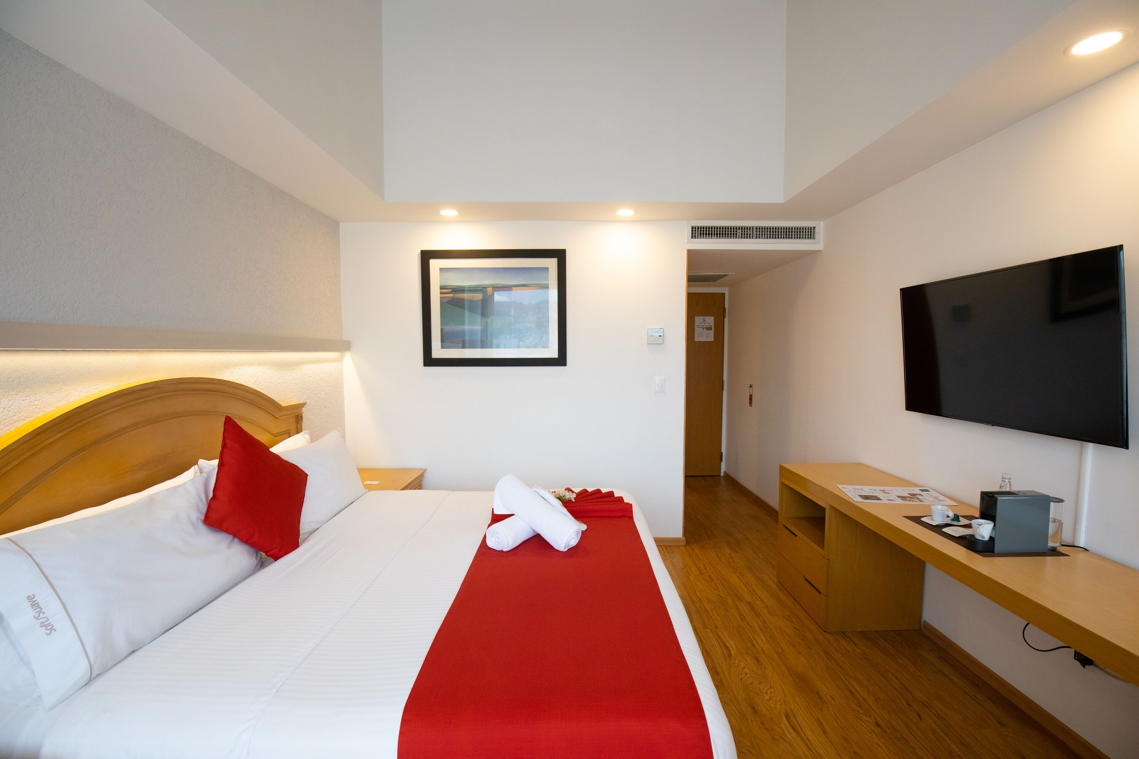 Quarto de hotel com uma cama, escrivaninha, televisão e um quadro na parede.