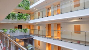 Interior de hotel com vários níveis, corrimãos de vidro e áreas verdes em sacadas.