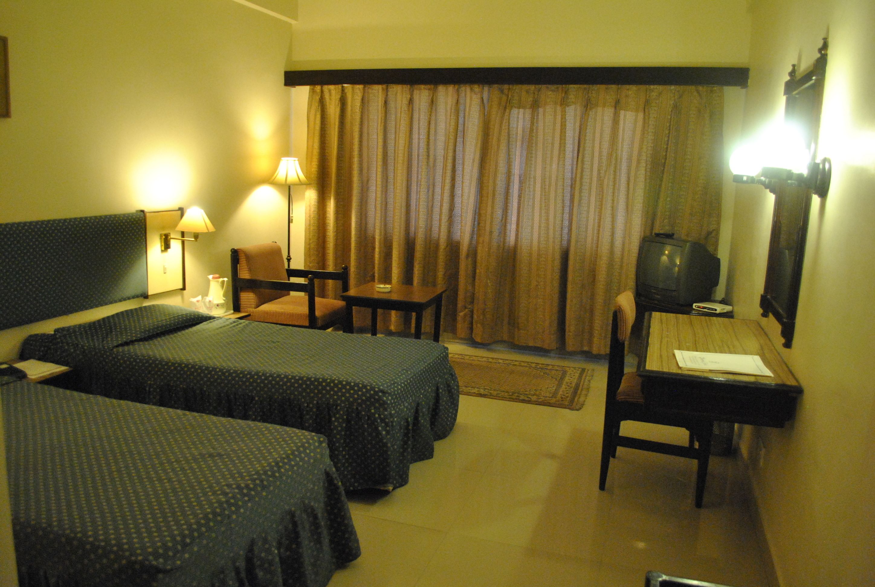 Quarto de hotel com duas camas, uma escrivaninha, uma cadeira, uma televisão e cortinas.