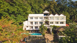 Um edifício branco de vários andares com piscina, rodeado por vegetação exuberante.
