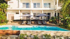 Uma área de piscina com espreguiçadeiras e guarda-sóis, um muro de pedra e um edifício com várias varandas.