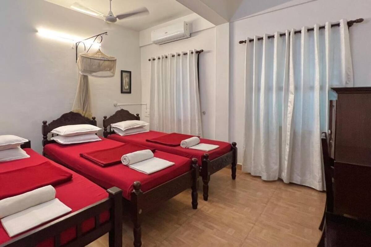 Quarto de hotel com três camas de madeira, cada uma com colcha vermelha e almofadas brancas. Há um ventilador de teto, um ar condicionado e uma janela com cortinas brancas.