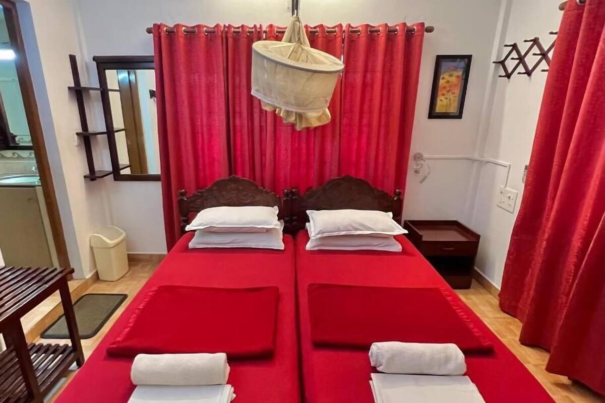 Quarto com duas camas individuais, cada uma com colcha vermelha e travesseiros brancos. À esquerda, um banco de madeira, uma pequena lixeira e uma mesa de cabeceira de madeira com abajur. Cortinas vermelhas e um quadro emoldurado na parede.