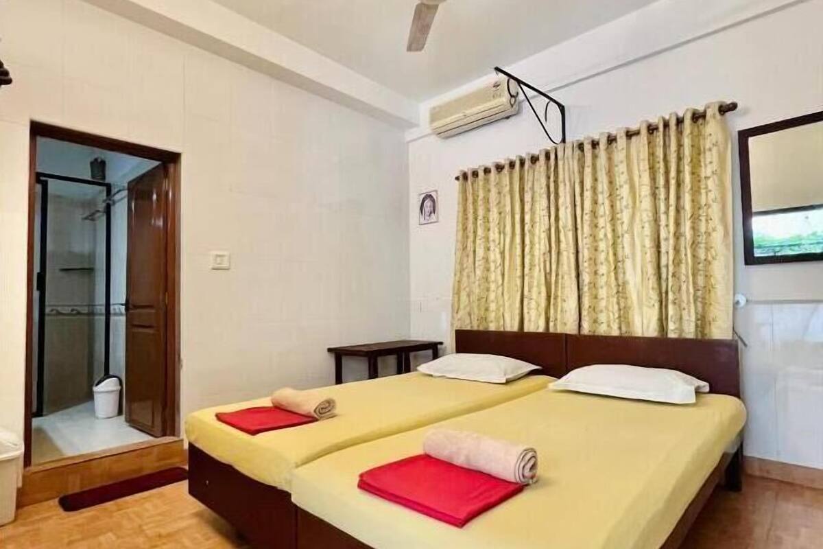 Uma cama de casal com duas toalhas vermelhas, um ventilador de teto, um aparelho de ar condicionado, piso de madeira e uma porta que dá para um banheiro.