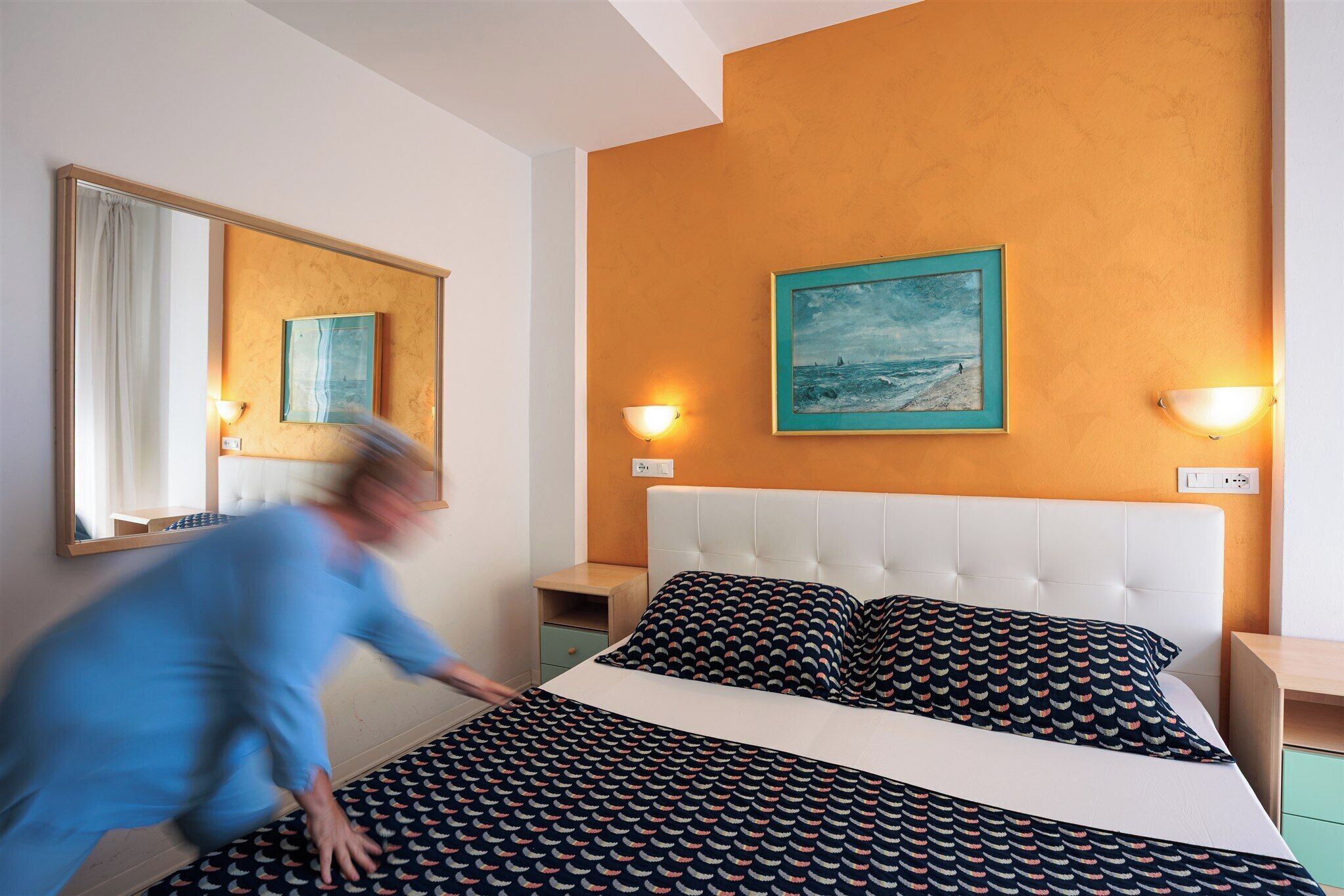 Quarto de hotel com cama, um espelho grande, um quadro e duas luzes fixadas na parede.