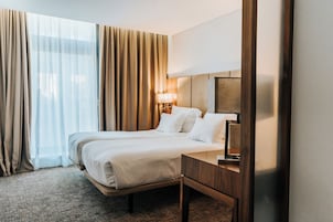 Quarto de hotel com cama, mesa de cabeceira, abajur e uma janela grande com cortinas.