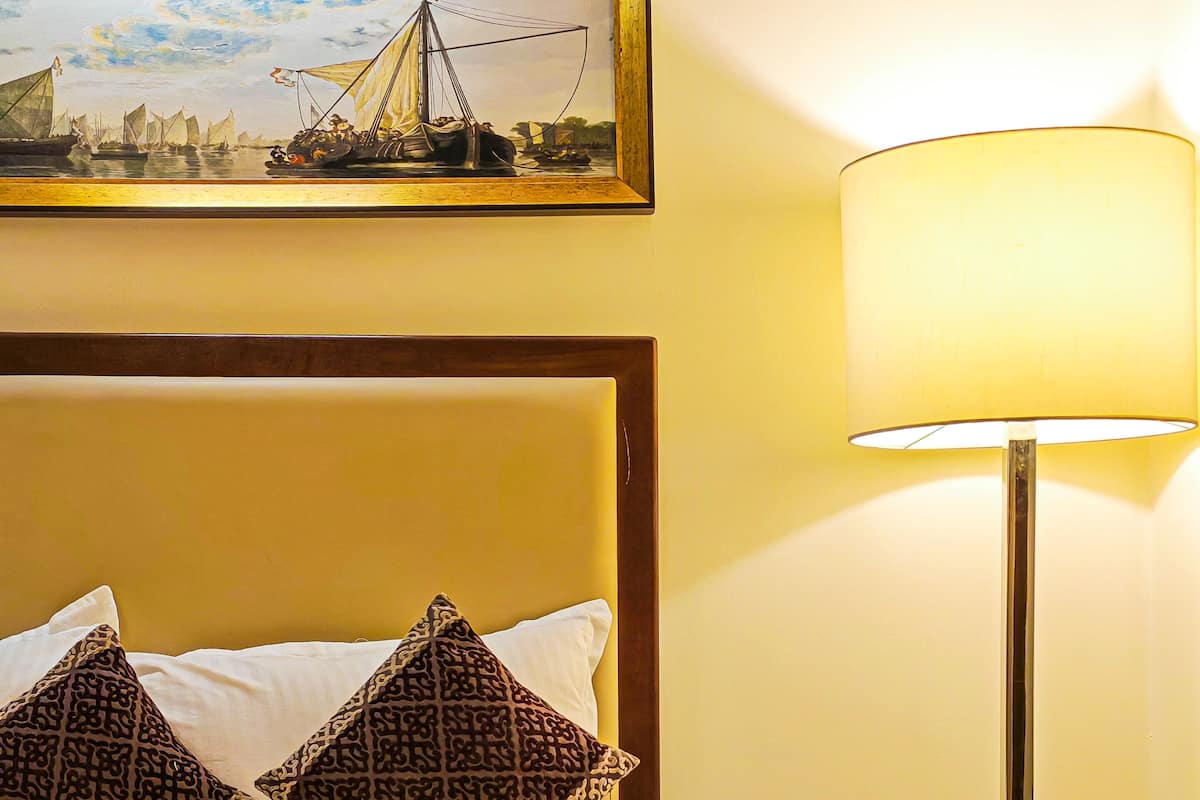 Cama bem arrumada com almofadas estampadas, um quadro emoldurado acima e um abajur de chão ao lado.