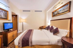 Quarto de hotel com uma cama, uma escrivaninha, uma televisão e um abajur.