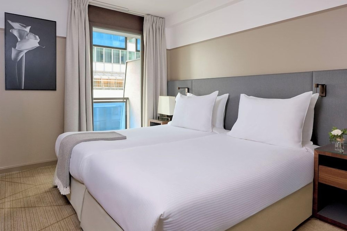 Quarto de hotel com uma cama grande, uma mesa de cabeceira, um abajur, um quadro na parede e vista para o exterior.
