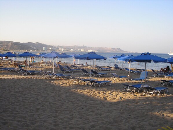 Uma praia com fileiras de espreguiçadeiras e guarda-sóis azuis, céu claro e vista distante de colinas e edifícios.