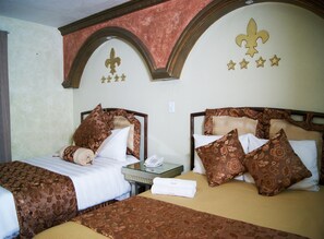 Quarto de hotel com duas camas, cada uma com roupa de cama e almofadas estampadas com flores, uma mesa de cabeceira com telefone, e uma parede decorada com motivos de lírio-fleur e estrelas.