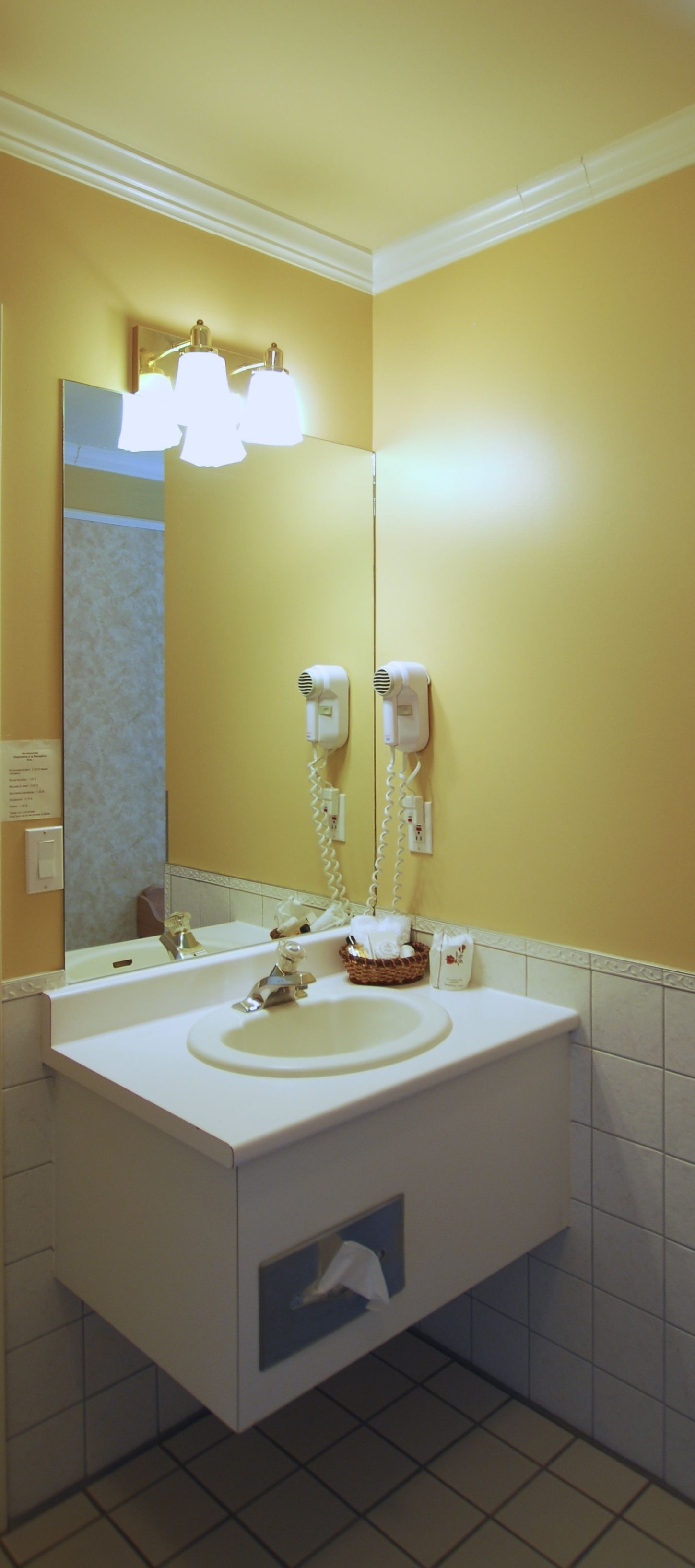 Casa de banho com lavatório branco, espelho e secador de cabelo fixado na parede.