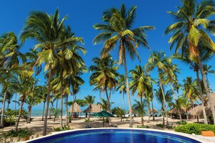 Vista da piscina com palmeiras, praia e céu azul límpido.