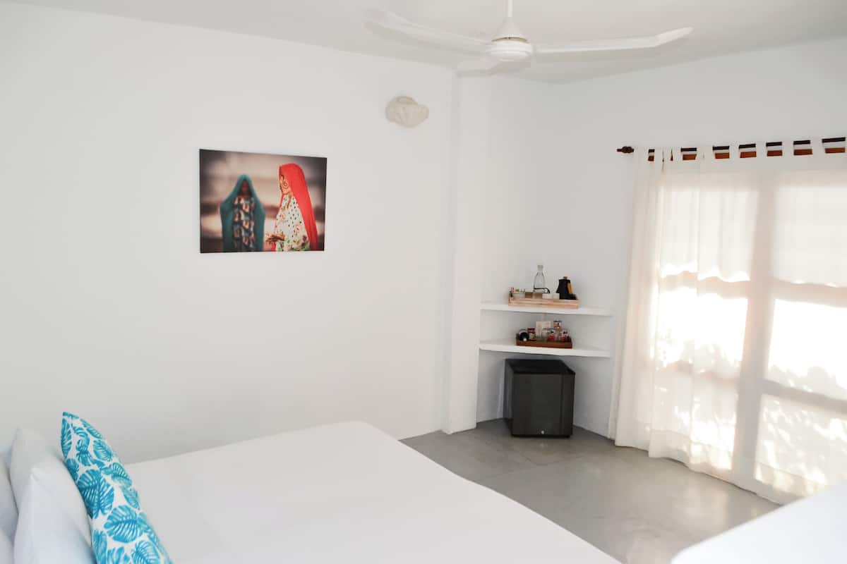 Quarto com uma cama, um ventilador de teto, uma prateleira pequena com objetos e uma janela com cortinas.