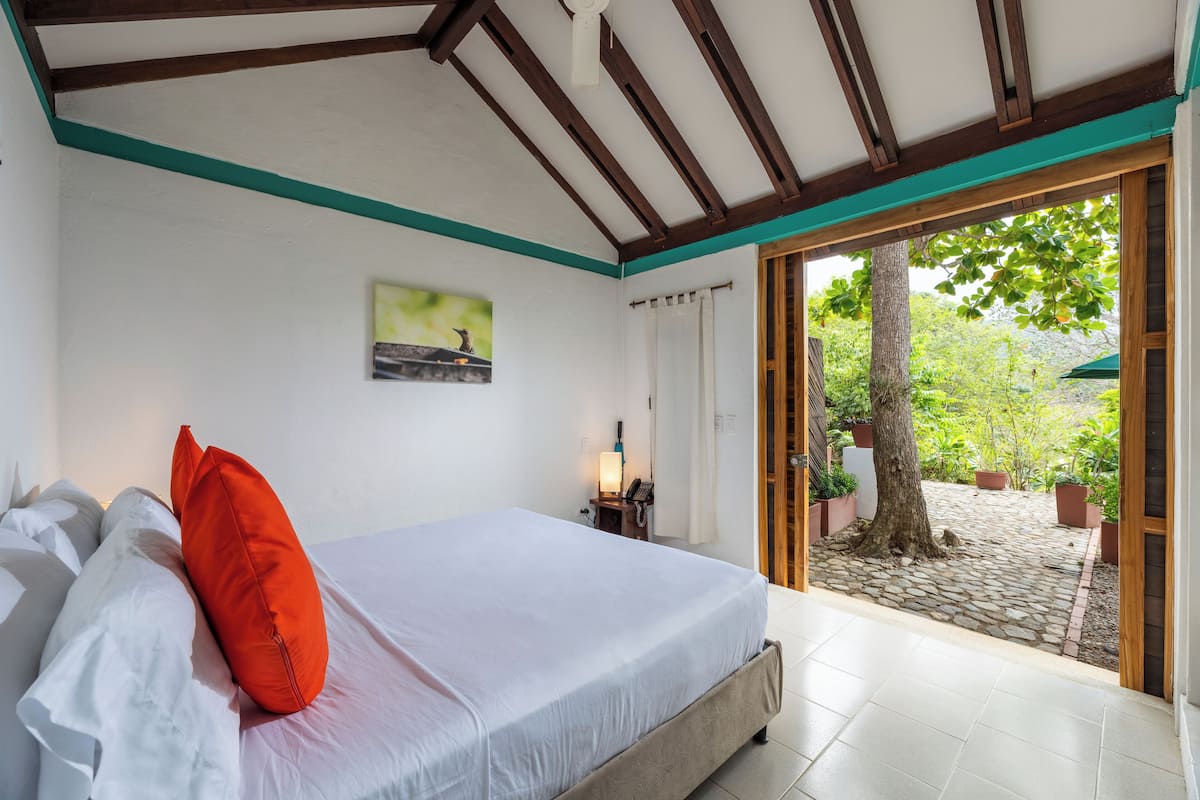 Quarto com cama, mesa de cabeceira, luminária e vista para um jardim através de uma porta aberta.