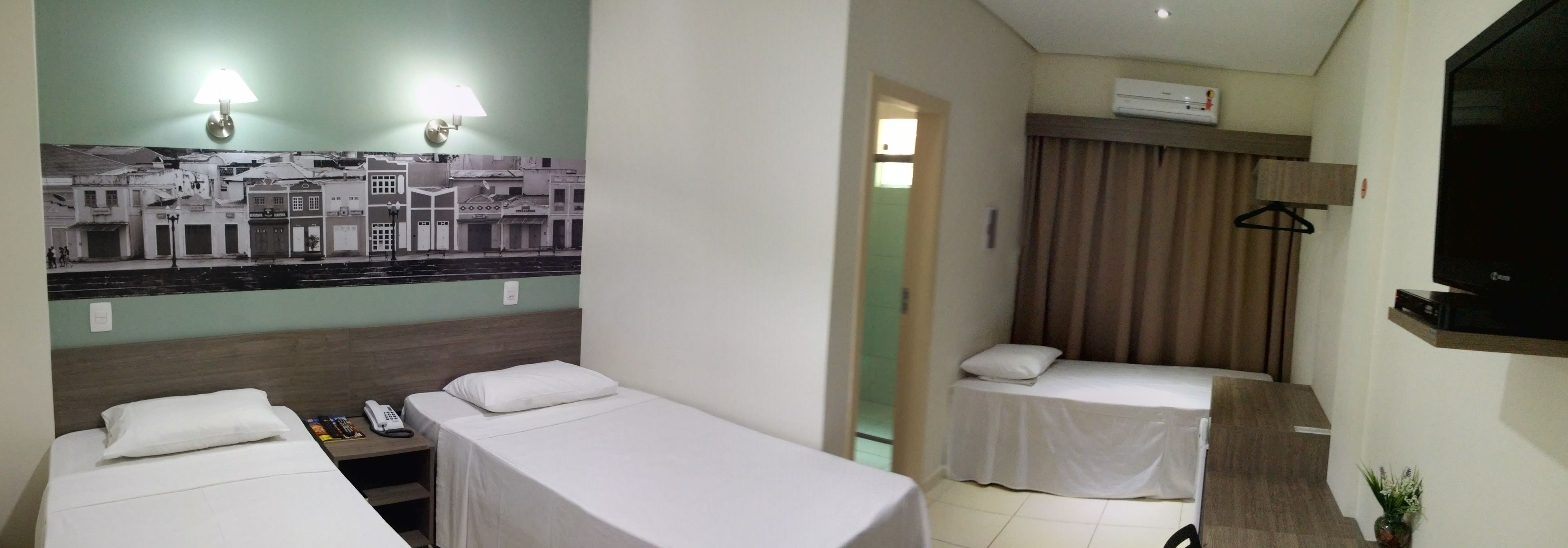 Quarto de hotel com duas camas de solteiro, uma mesinha com telefone, televisão na parede e um quadro com vista urbana.