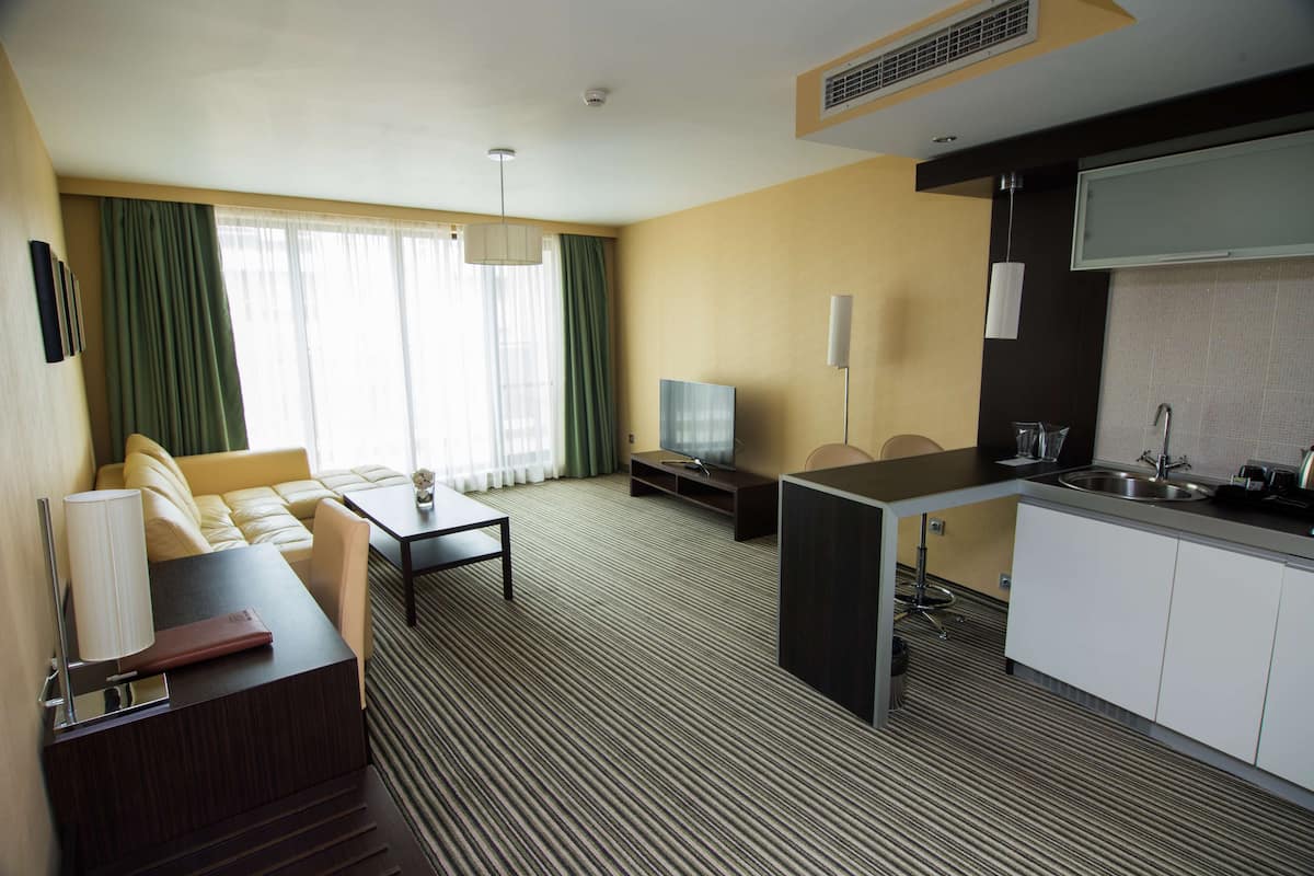 Quarto de hotel com sofá, mesa de centro, televisão, pequeno-almoço e uma janela grande com cortinas.