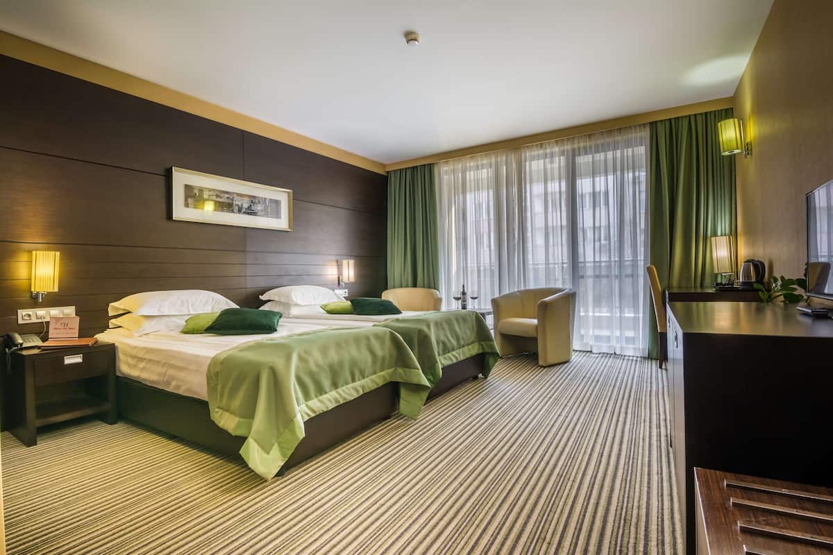 Quarto de hotel com duas camas, uma escrivaninha, uma cadeira, uma mesa de cabeceira, uma luminária e um quadro na parede.