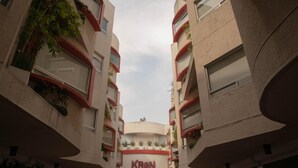 Um edifício com vários andares, um átrio central, varandas com plantas e uma placa 'KRON' bem visível.
