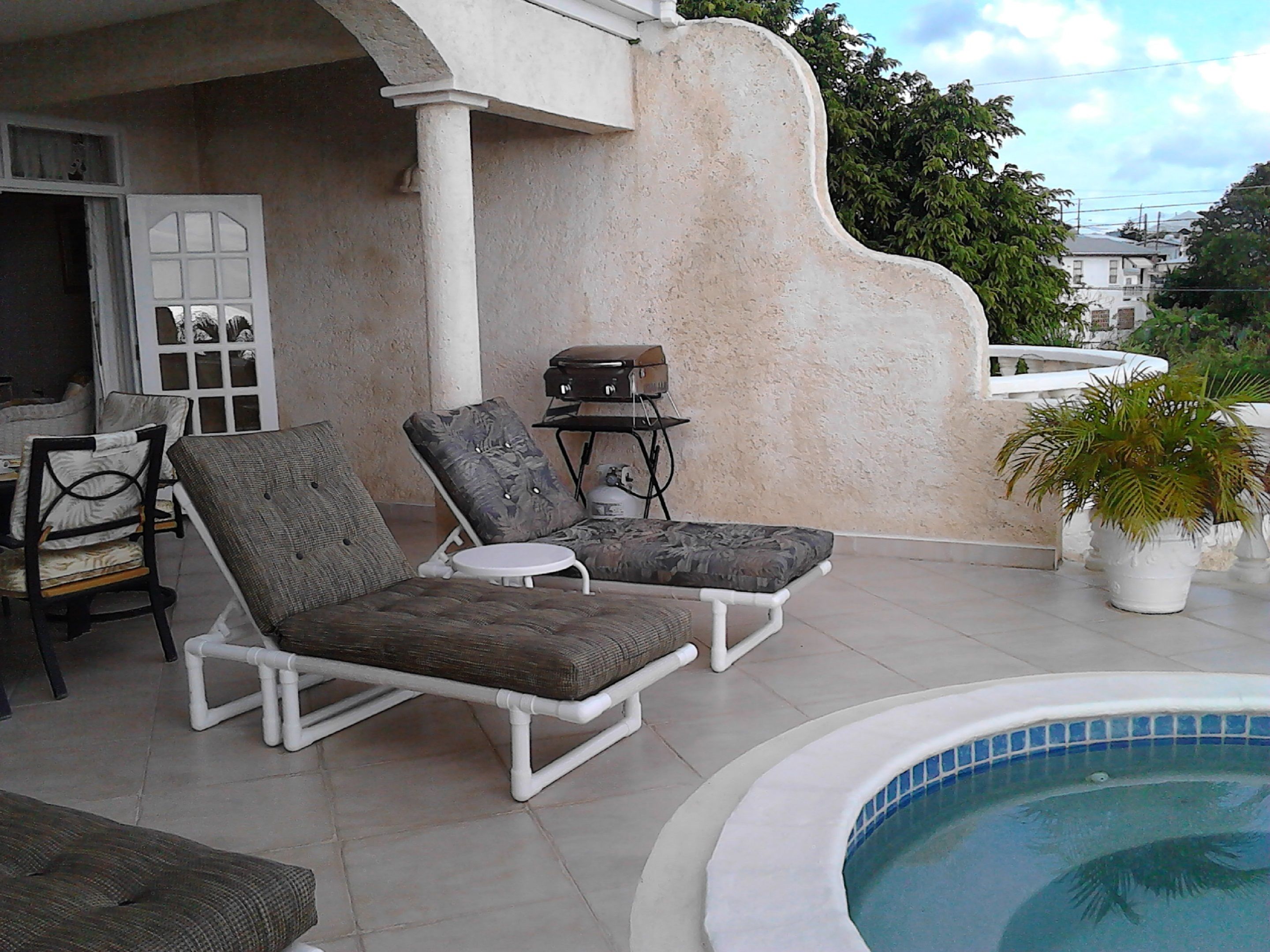 Uma área à beira da piscina com espreguiçadeiras, uma mesa pequena e um vaso com planta.
