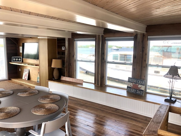 Um quarto aconchegante com piso de madeira, uma mesa redonda com jogos americanos trançados, um assento junto à janela com vista para a água, e uma placa com a inscrição 'SANDY TOES AND SALTY KISSES'.