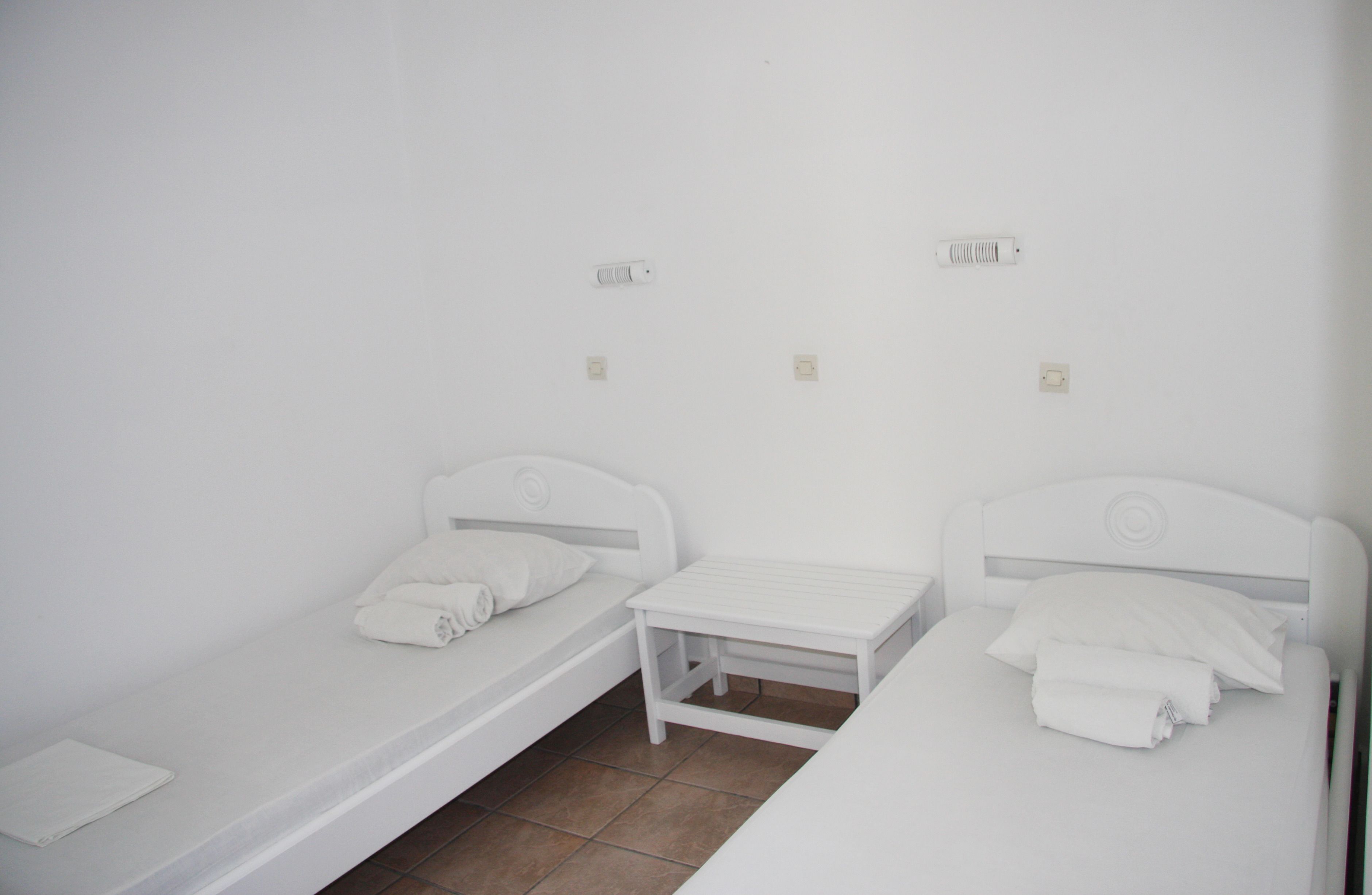 Quarto com duas camas de solteiro, cada uma com cabeceira branca e uma mesinha de cabeceira.