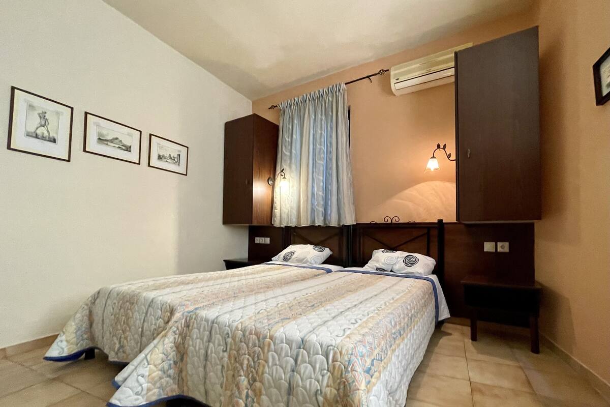 Uma cama de casal com colcha estampada, mesa de cabeceira, guarda-roupa e ar condicionado.