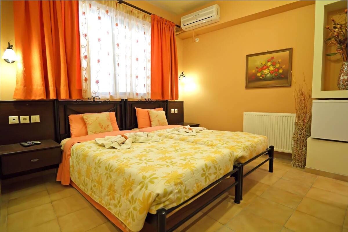Quarto com cama, mesas de cabeceira, ar condicionado fixado na parede, um quadro e um vaso com flores.