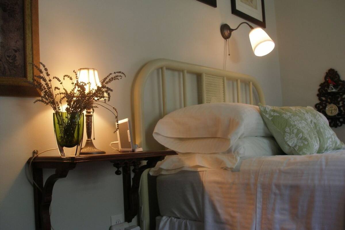 Quarto com cama, mesa de cabeceira, luminária e relógio fixado na parede.
