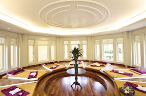Um quarto circular com macas de massagem dispostas em torno de uma mesa central, janelas amplas e teto decorativo.