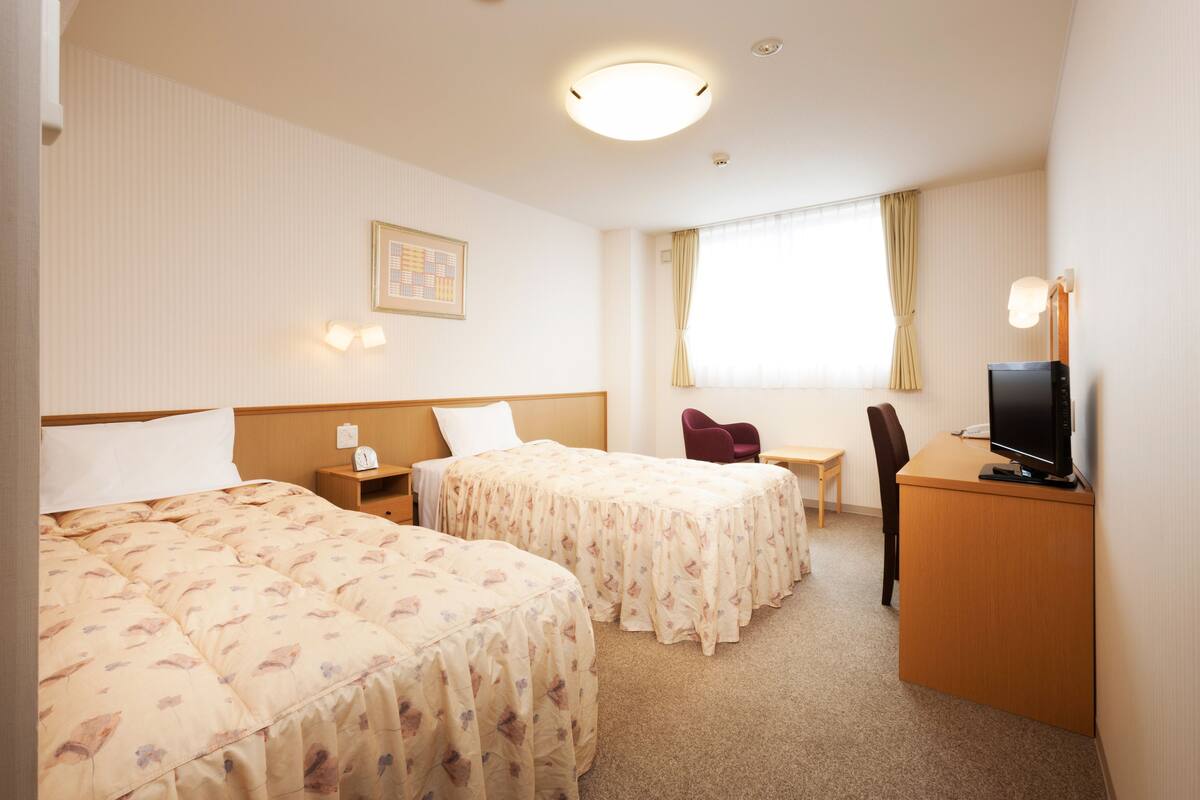Quarto de hotel com duas camas, uma escrivaninha com computador, uma cadeira e uma televisão.