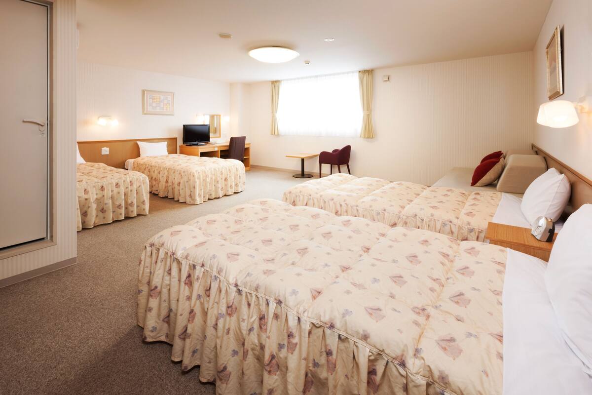Quarto de hotel com duas camas, uma mesa, uma cadeira, uma televisão e uma janela com cortinas.