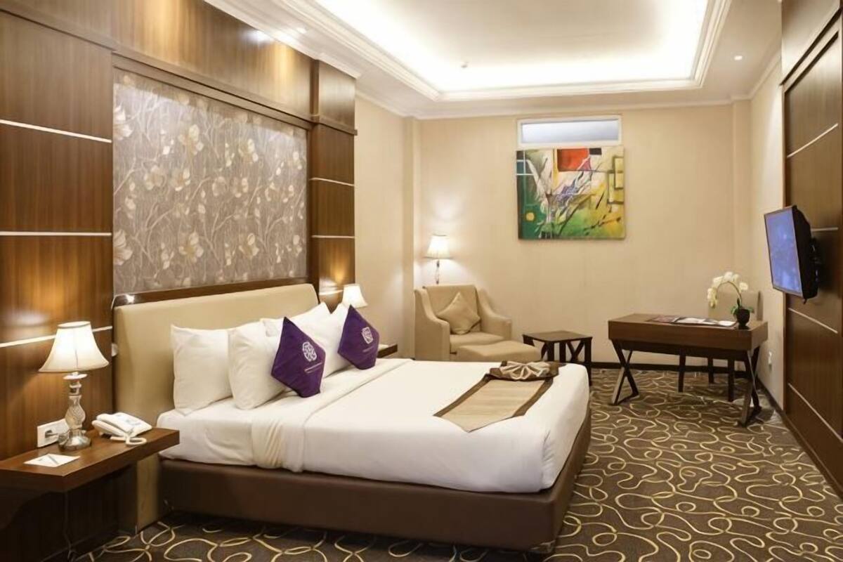 Quarto de hotel com cama, escrivaninha, cadeira, televisão e um quadro na parede.