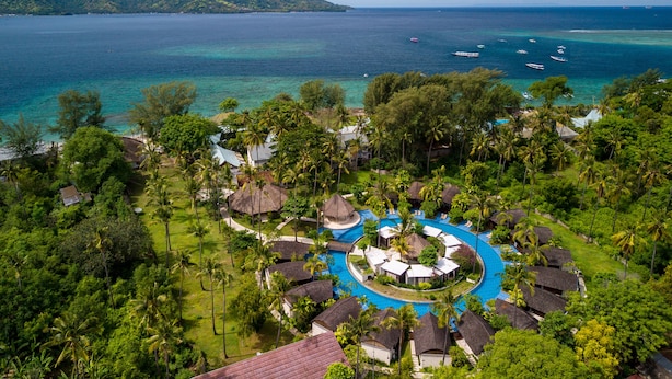 Vista aérea de um resort com uma piscina circular rodeada por acomodações tipo bangalô, com o mar azul cristalino e uma cordilheira distante ao fundo.