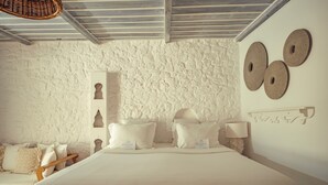 Quarto com parede em textura de pedra, cama com roupa de cama branca, um banco de madeira e uma prateleira com objetos decorativos.
