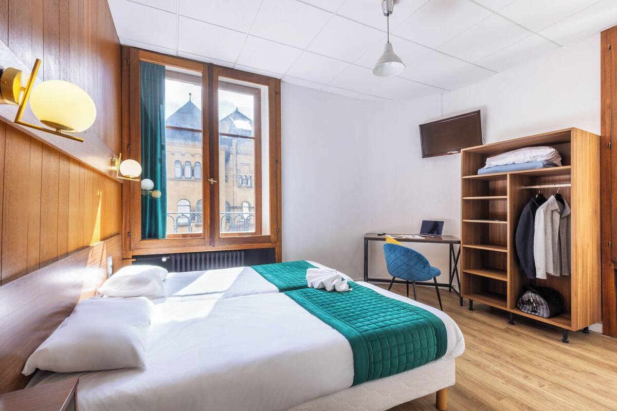Quarto de hotel com cama, escrivaninha, cadeira, guarda-roupa e janela com vista para edifícios.