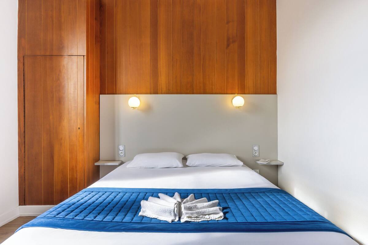 Quarto de hotel com cabeceira de madeira, duas luminárias de apoio, cama com roupa de cama branca e almofada azul decorativa.
