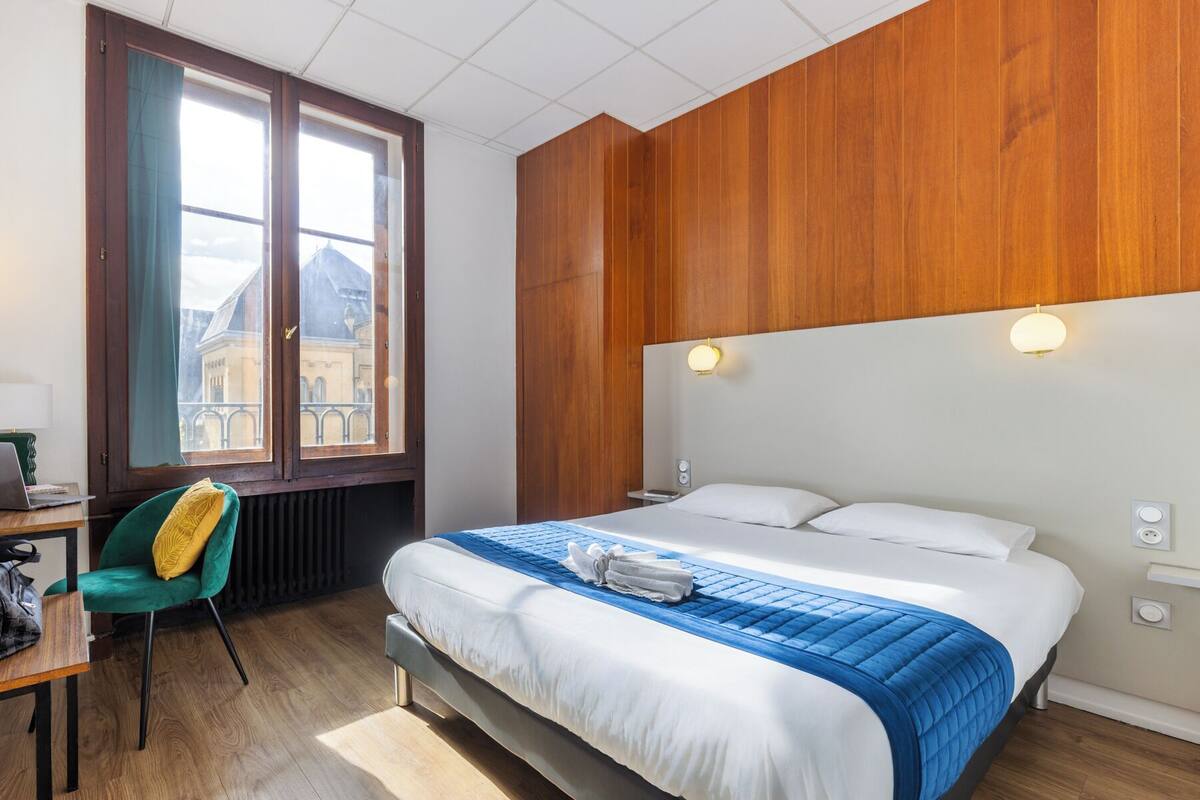 Quarto de hotel com uma cama, uma escrivaninha com um laptop, uma cadeira e uma janela com cortinas.