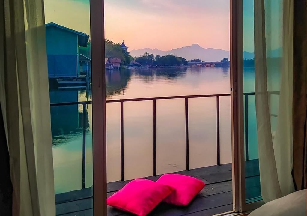 Varanda com vista para um lago tranquilo e montanhas ao longe, com grade e almofadas no chão.